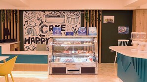 Um balcão de pastelaria com uma variedade de produtos de panificação expostos em uma vitrine de vidro, um menu em lousa e uma parede com um mural com tema de bolo.