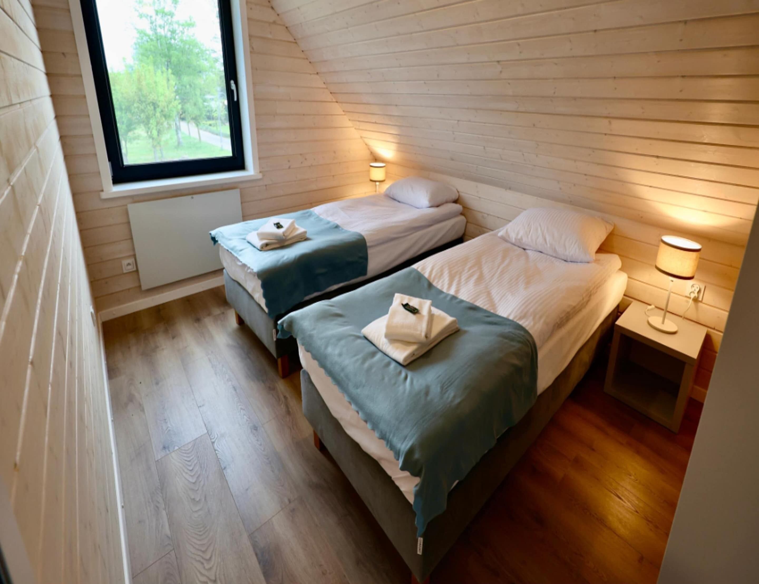 Um quarto pequeno e acolhedor, com duas camas de solteiro, uma janela e uma mesa de cabeceira com abajur.
