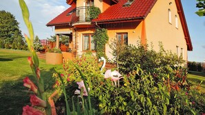 Uma casa de dois andares com telhado de telhas vermelhas, varanda e um jardim com diversas plantas e flores.