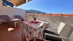 Um terraço com duas cadeiras de estar, uma mesa com toalha de mesa florida e um vaso com flores.