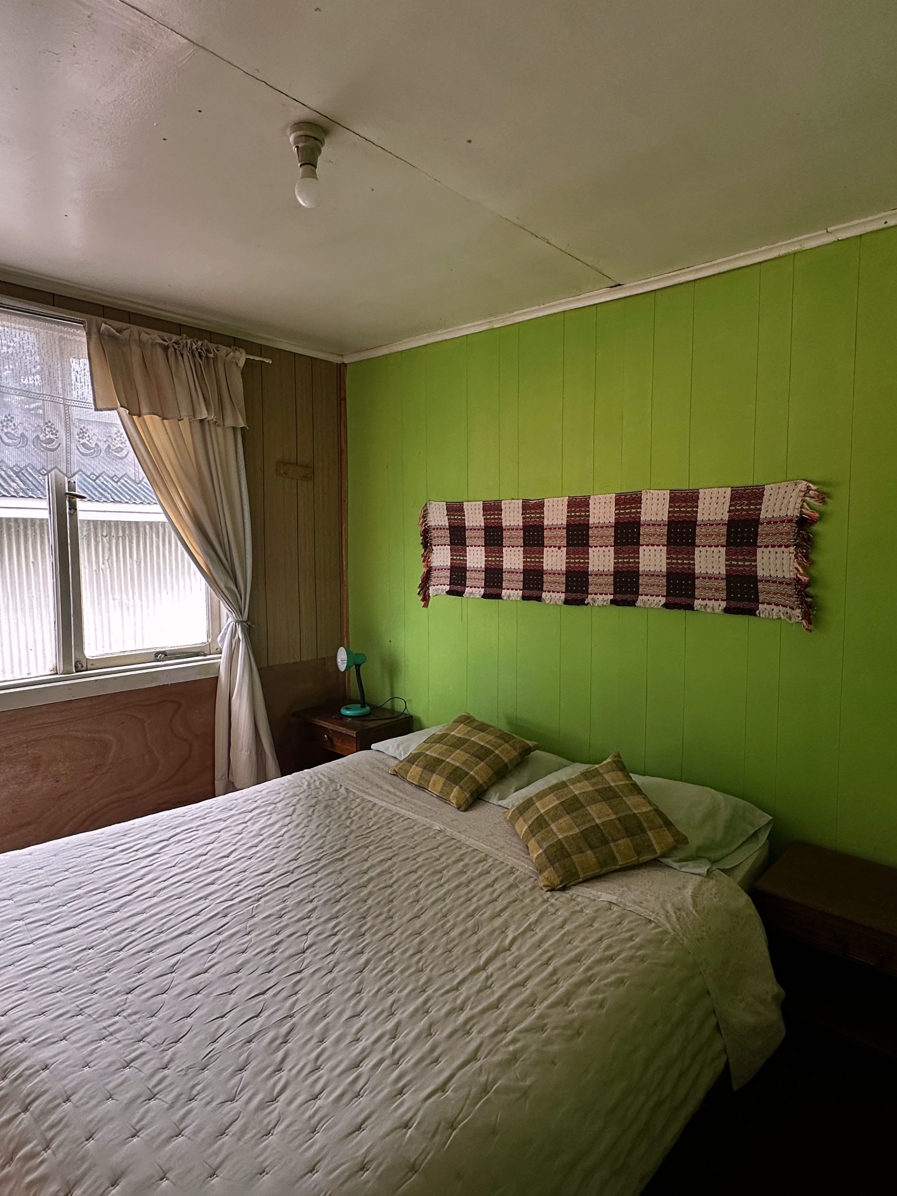 Quarto com cama, manta na parede, mesa de cabeceira com abajur e janela com cortinas.