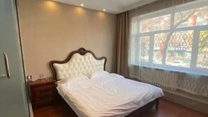 Quarto com cama grande, mesa de cabeceira de madeira e janela com vista para árvores e edifícios.