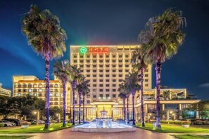 Um hotel com uma fonte central, rodeado por palmeiras e uma entrada bem iluminada.