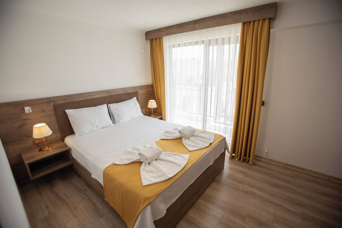 Quarto de hotel com cabeceira de madeira, uma cama grande com roupa de cama branca, duas luminárias de cabeceira e uma janela com cortinas.