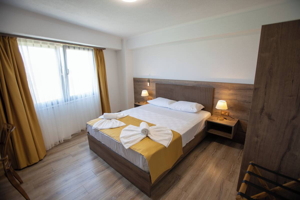 Quarto de hotel com cama de madeira, duas mesas de cabeceira com luminárias, guarda-roupa de madeira e janela com cortinas.