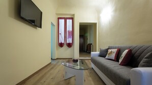 Uma sala de estar com uma televisão de tela plana na parede, uma mesa de centro de vidro com uma tigela de frutas, um sofá cinza com almofadas decorativas e um piso de madeira.