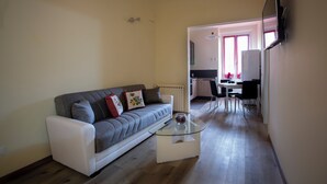 Uma sala de estar com um sofá, uma mesa de centro de vidro e uma TV fixada na parede.