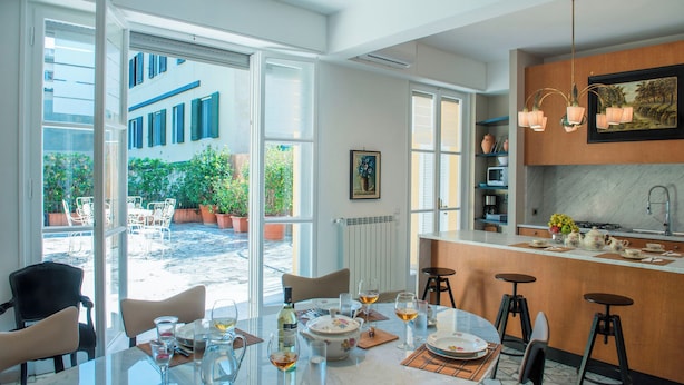 Uma sala de jantar com mesa posta para quatro, cozinha com bancada de mármore e vista para um edifício e vegetação.