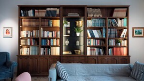 Quarto com uma grande estante de madeira cheia de livros, uma poltrona azul, uma cadeira rosa e uma cama com roupa de cama listrada.
