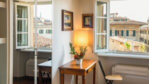 Quarto de canto com uma mesa de madeira, uma cadeira, um abajur e uma janela com vista para edifícios.