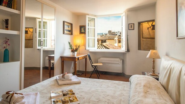 Quarto com cama, escrivaninha, cadeira, janela com vista para prédios e estante de livros.