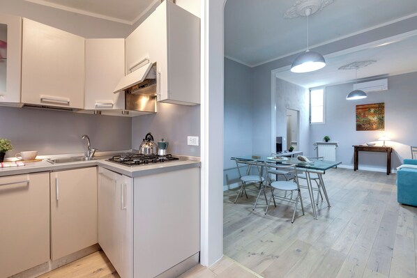 Cozinha moderna com armários brancos e fogão a gás, sala de jantar com mesa de vidro e cadeiras de metal, e sala de estar com sofá azul e mesa de centro de madeira.