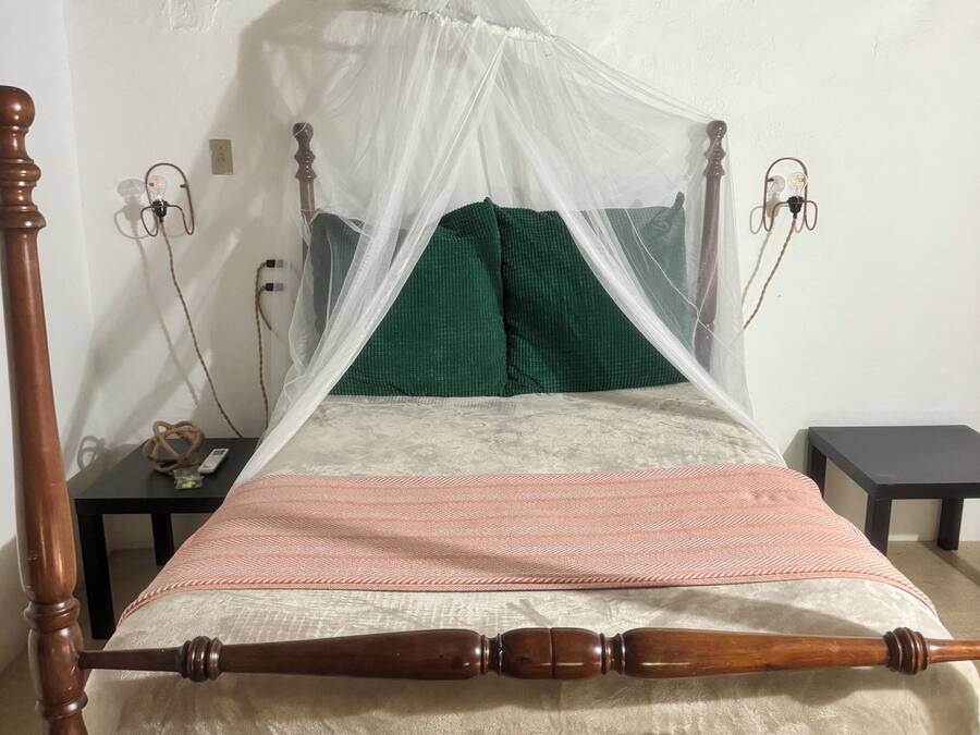 Uma cama de madeira com dossel, almofadas verdes e um edredom listrado em rosa e branco. Há mesas de cabeceira com abajures e uma tomada na parede.