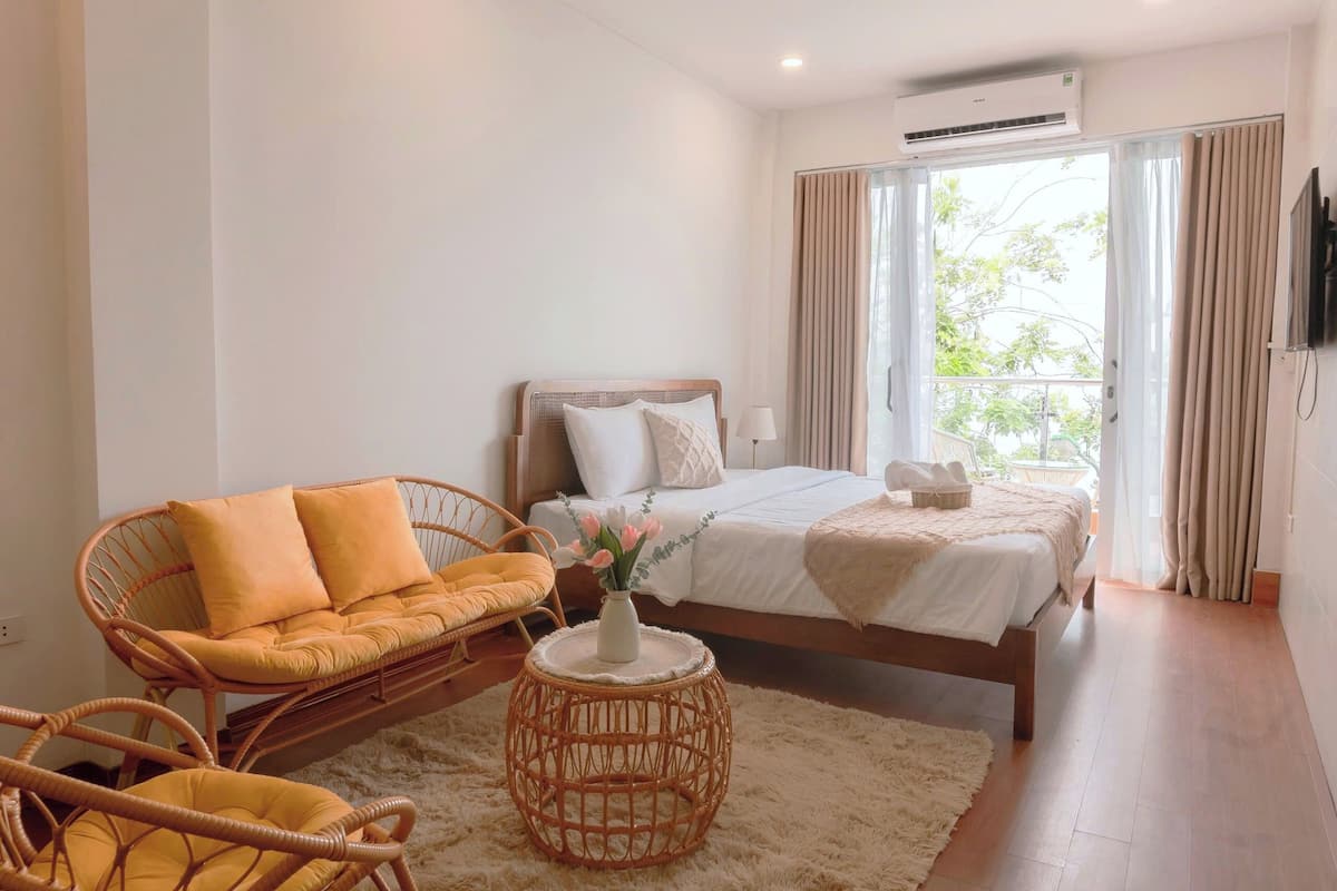 Um quarto com uma cama, uma cadeira de vime, uma mesa redonda pequena com um vaso, e vista para o exterior pela janela.