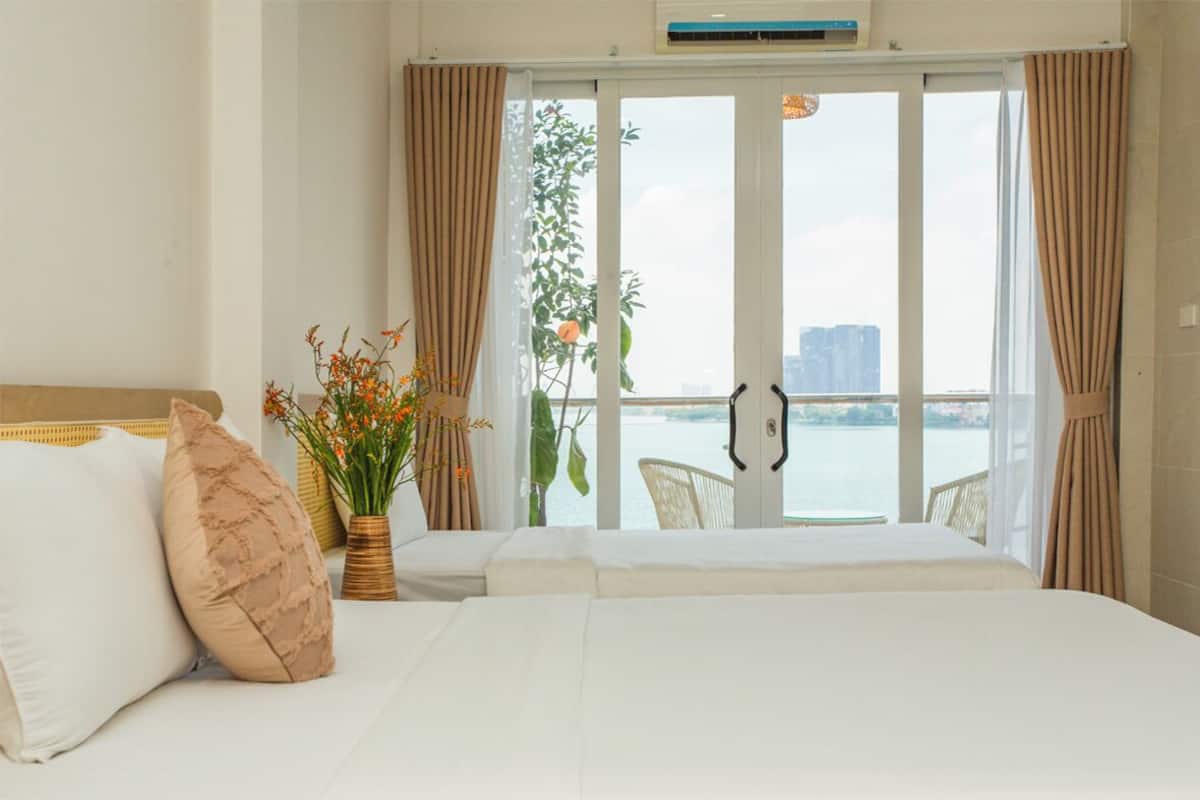 Quarto de hotel com cama, janela com cortinas, vista para a água e uma planta no parapeito.