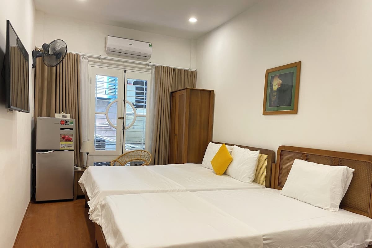 Quarto de hotel com duas camas, um frigorífico, ar condicionado, janela com cortinas, uma cadeira e um quadro na parede.