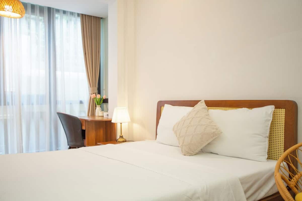 Quarto de hotel com cama, escrivaninha com luminária, cadeira e janela com cortinas.