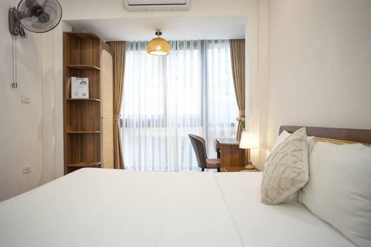 Quarto de hotel com cama, escrivaninha, cadeira, ventilador e janela com cortinas.
