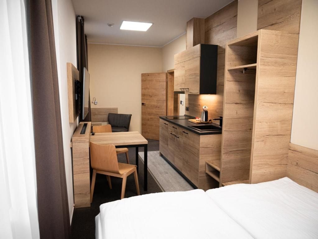 Quarto de hotel pequeno com cama, escrivaninha, cadeira e cozinha compacta.