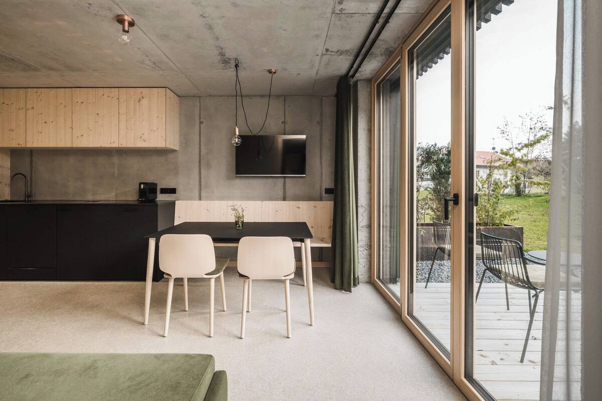 Cozinha moderna com armários de madeira, bancada preta e televisão montada na parede. Há um espaço de jantar com mesa e duas cadeiras, e uma grande porta de vidro deslizante que dá para um pátio externo com mesa e cadeiras.