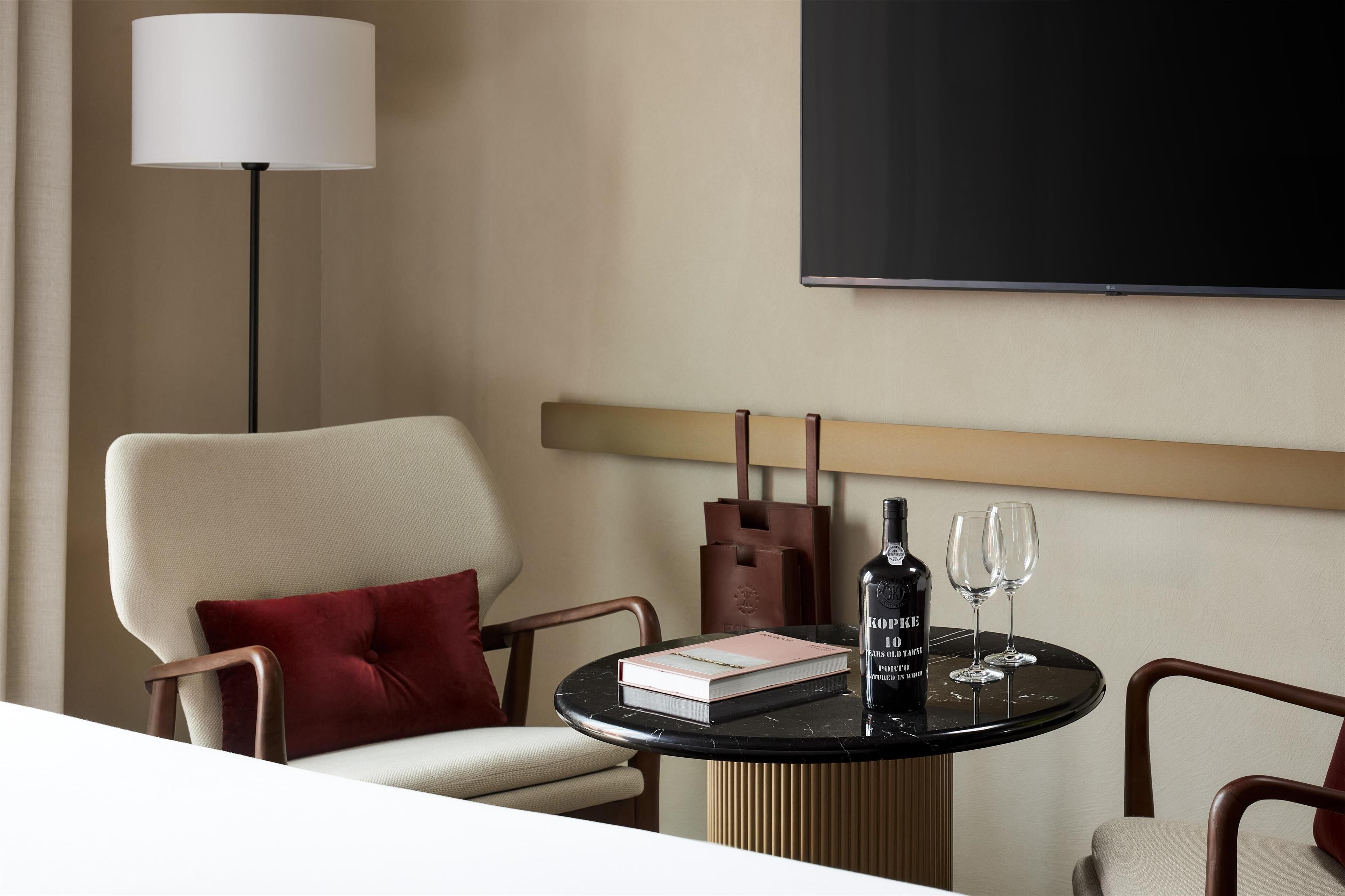 Quarto de hotel com design moderno, apresentando uma grande televisão de tela plana na parede, uma mesa redonda preta com uma garrafa de vinho e copos, uma poltrona confortável com almofada vermelha e uma estante com um livro.
