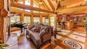 Interior de cabana de tronco com um piano de cauda, um sofá estampado e uma área de jantar com balcão.