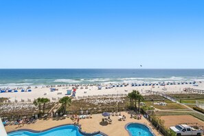 Vista da praia a partir de uma varanda de hotel, com piscina, espreguiçadeiras e uma área de areia.