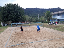 Uma quadra de vôlei de areia com rede, rodeada por árvores e um edifício com fachada azul.