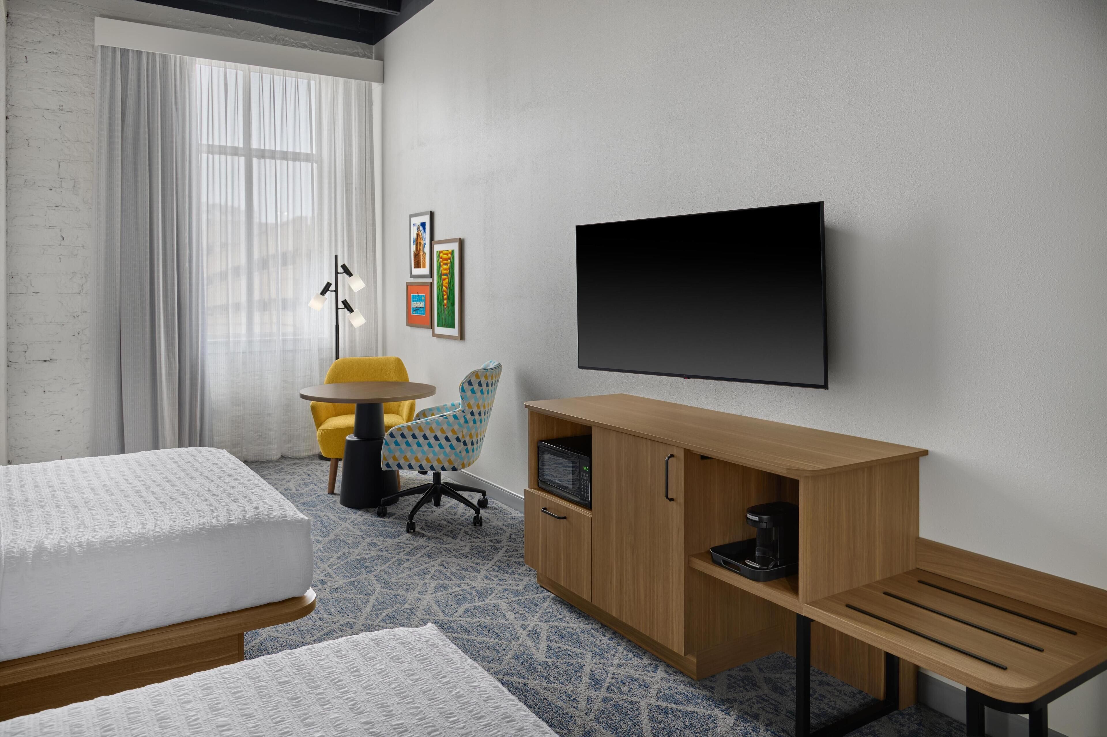 Quarto de hotel com uma cama, escrivaninha, cadeira, televisão e obras de arte na parede.