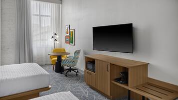 Quarto de hotel com uma cama, escrivaninha, cadeira, televisão e obras de arte na parede.