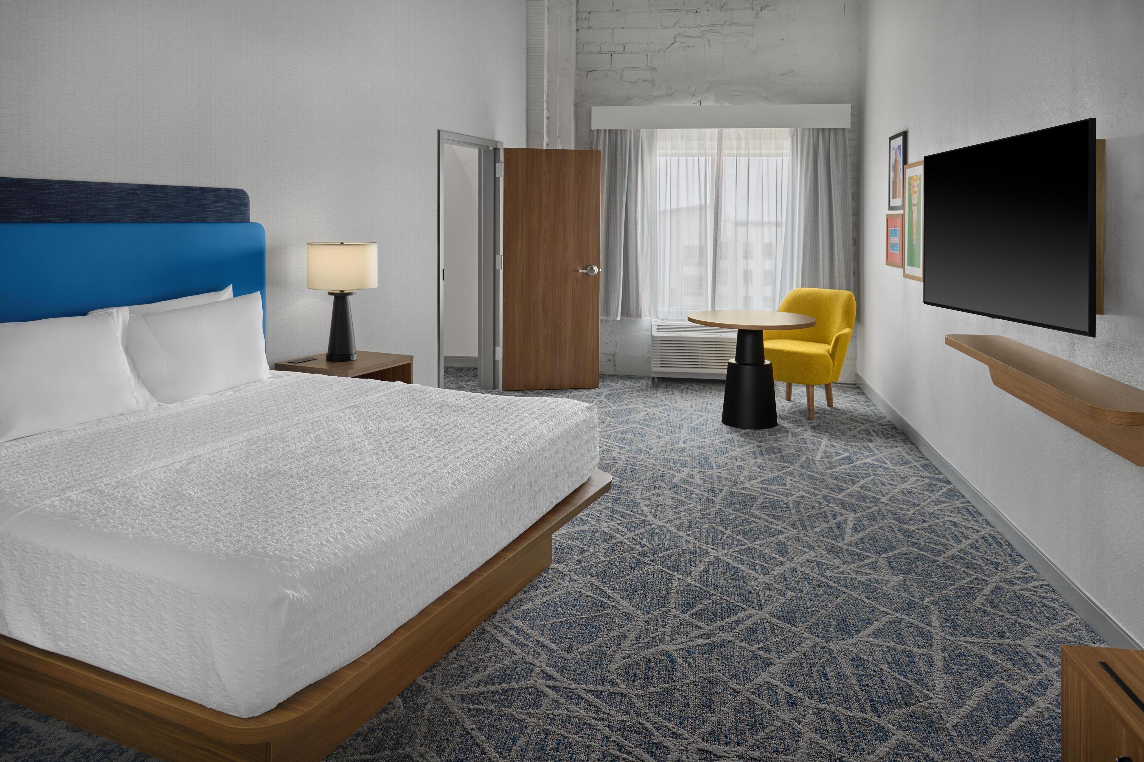 Quarto de hotel com uma cama grande, uma mesa com luminária, uma cadeira, uma televisão e uma janela com cortinas.