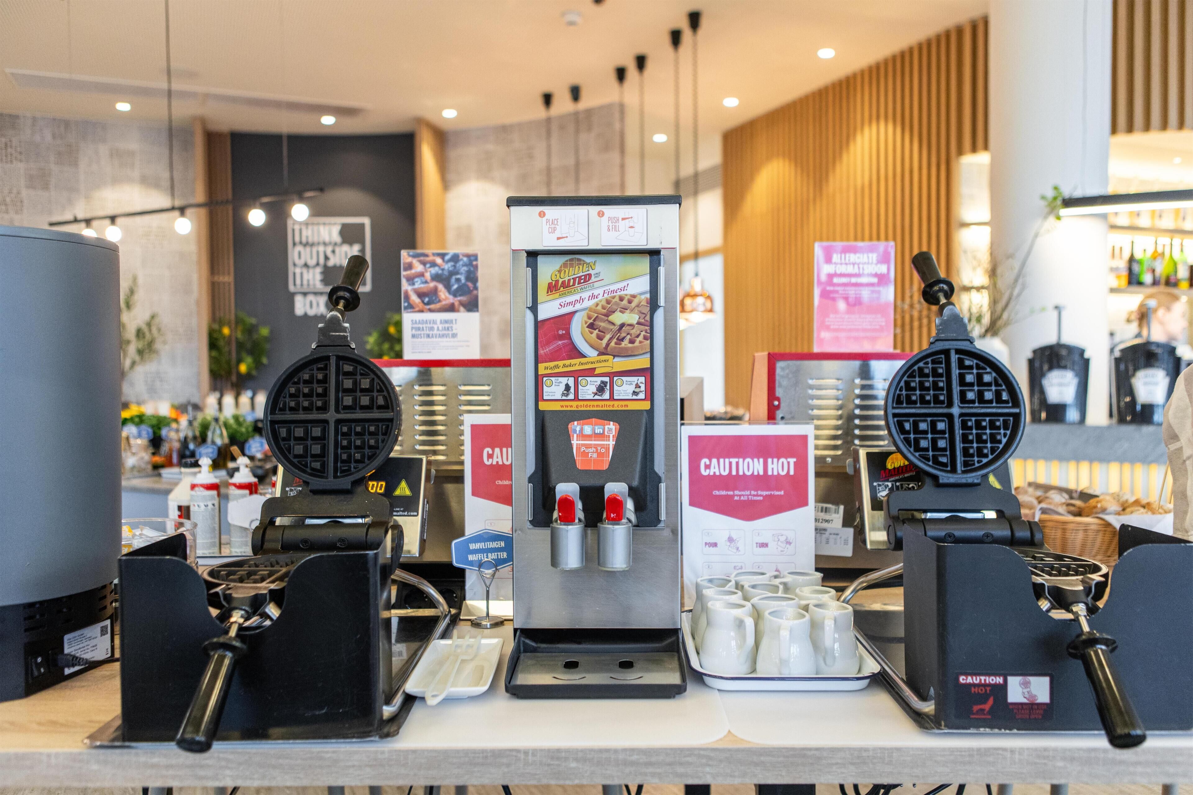 Uma estação de waffles com duas máquinas de waffle e uma prensa, um aviso de precaução em placa, e um quadro de menu em um café.