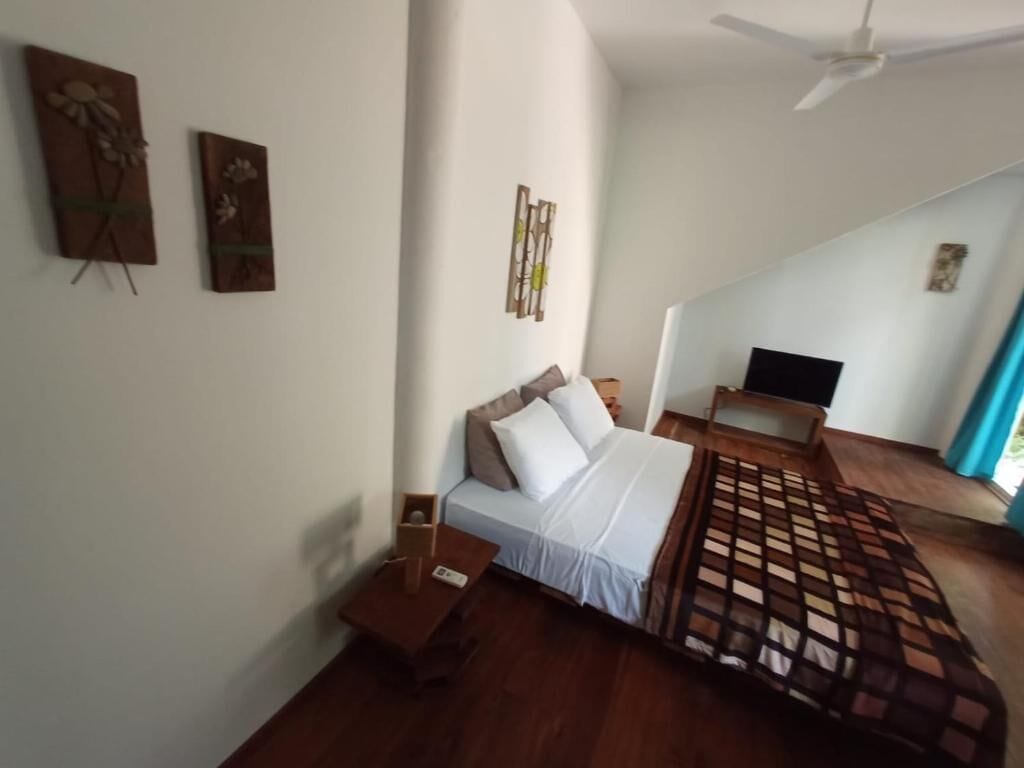 Quarto com cama, mesa de cabeceira e televisão. Há uma janela com cortinas e um ventilador de teto.