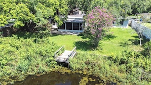 Uma casa com varanda coberta, um pequeno píer sobre um lago e vegetação exuberante.