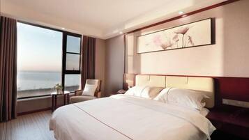 Quarto de hotel com uma cama grande, uma cadeira, uma mesinha de cabeceira, um quadro na parede e vista para o mar através de uma janela ampla.