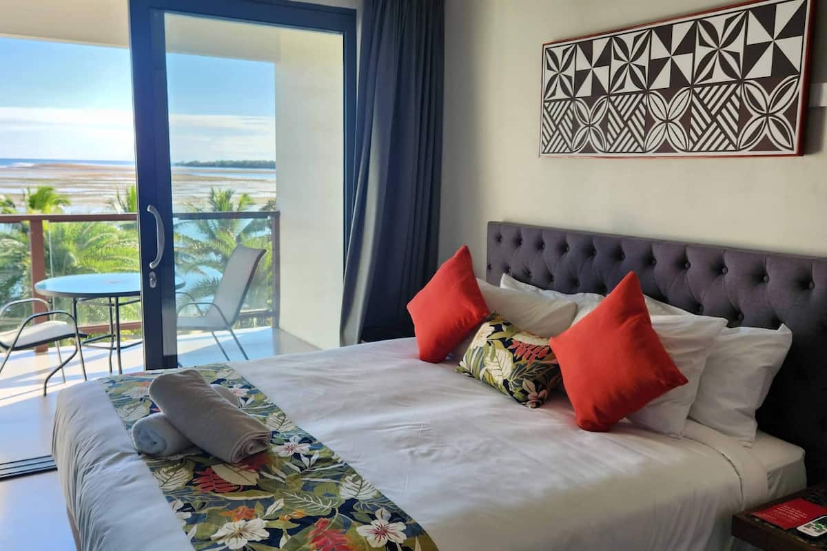Quarto de hotel com uma cama grande, vista para a praia, varanda com cadeiras e um ornamento geométrico na parede.