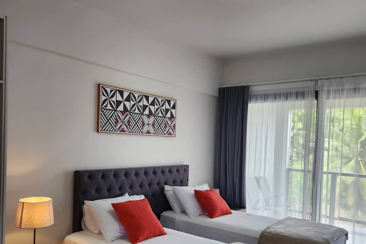 Quarto de hotel com duas camas, uma mesa de cabeceira, uma luminária, um objeto decorativo na parede e uma janela com cortinas.