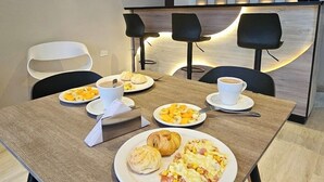 Uma área de refeições moderna com mesa de madeira posta para o café da manhã, incluindo croissants, ovos e frutas. Há cadeiras pretas e um balcão com um detalhe iluminado na parede ao fundo.