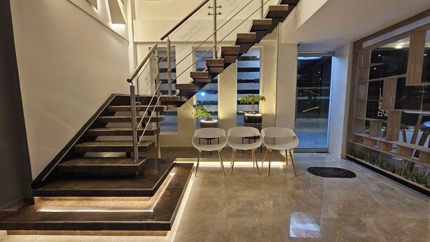 Escadaria moderna com guarda-corrimão de vidro, que leva a uma área de estar com três cadeiras brancas e uma mesa pequena.