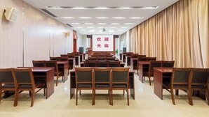 Uma sala de conferências com fileiras de cadeiras e mesas de madeira, teto com iluminação embutida e um banner com caracteres chineses.