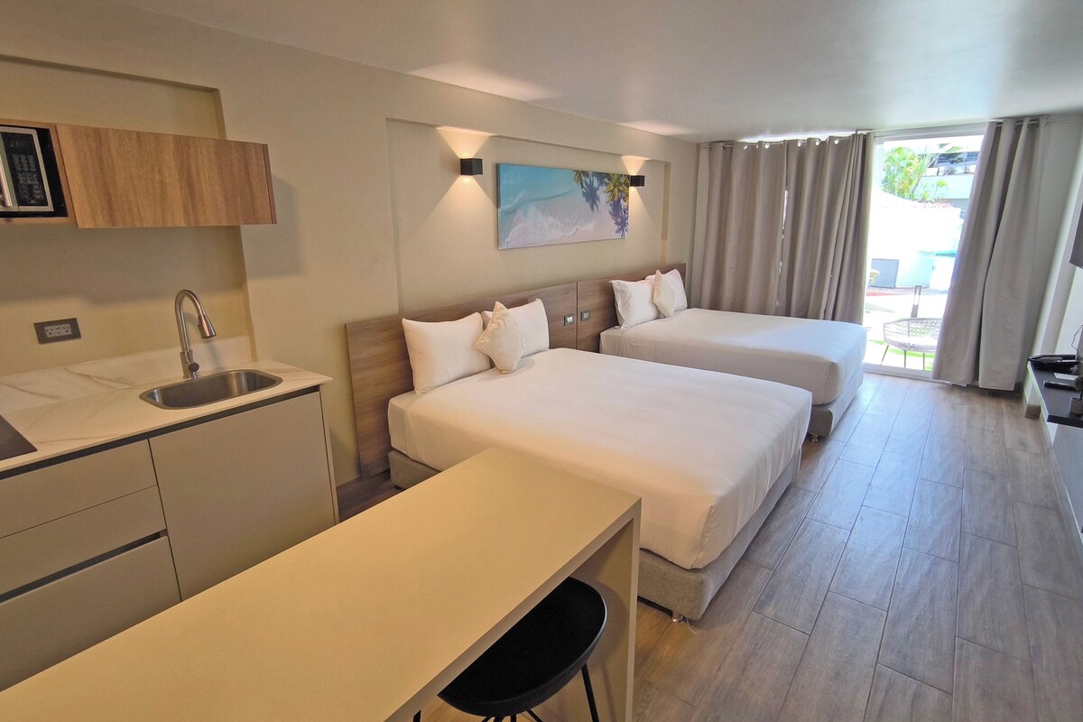 Quarto de hotel com cozinha compacta, duas camas, um quadro e uma televisão.