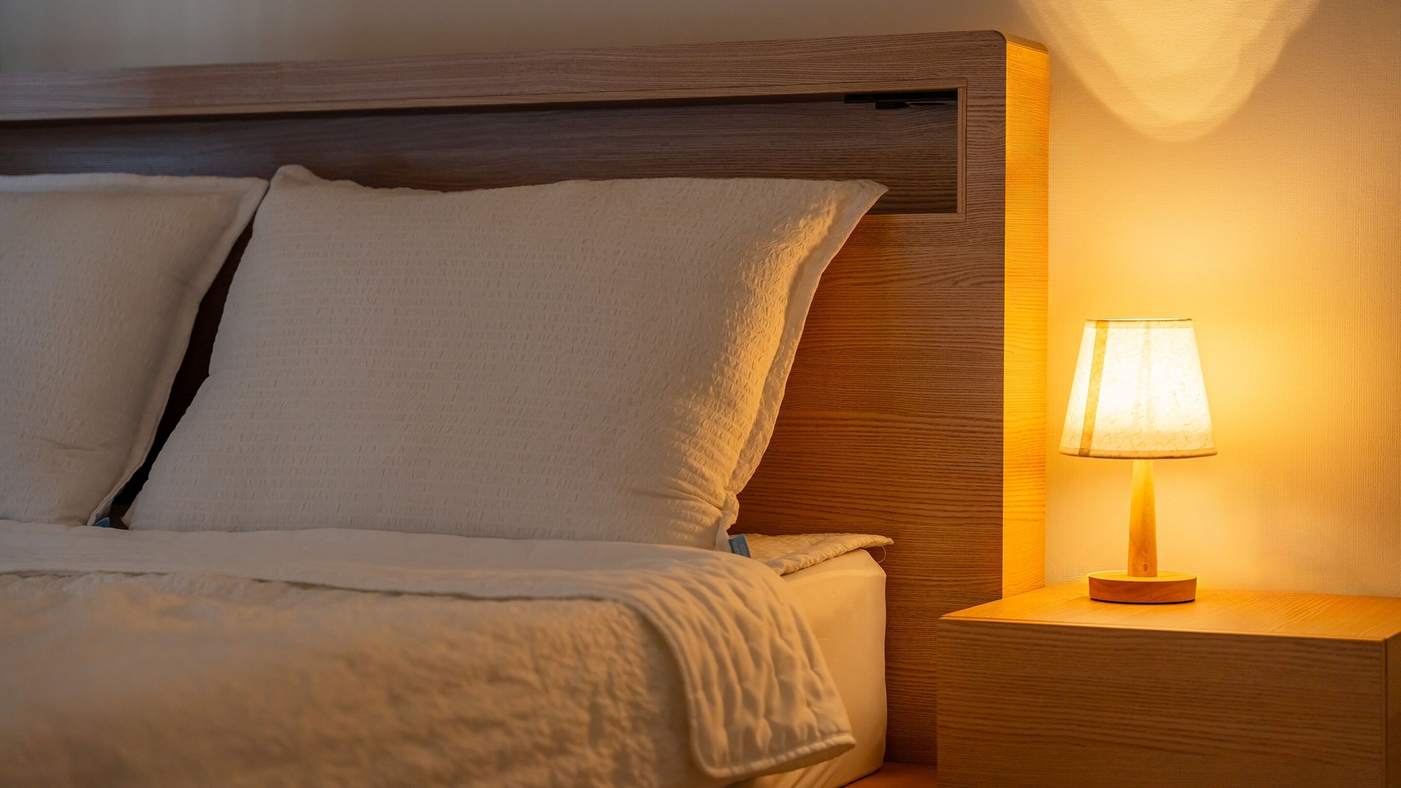 Uma cama bem arrumada com lençóis e almofadas brancas, uma cabeceira de madeira e uma mesa de cabeceira com abajur.
