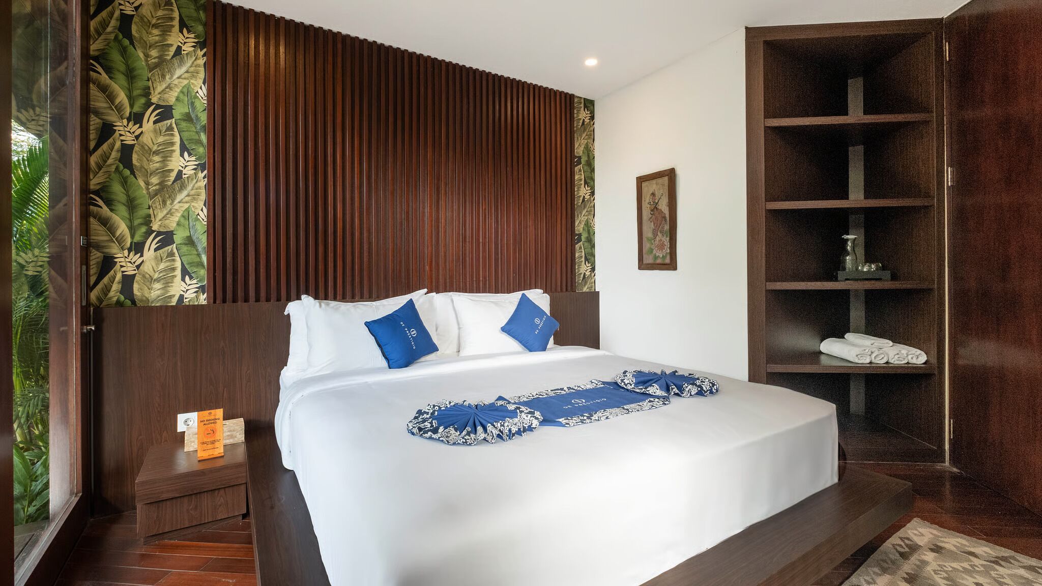 Cama bem arrumada com roupa de cama branca e azul, cabeceira de madeira e mesa de cabeceira com um telefone e uma caixa laranja pequena.