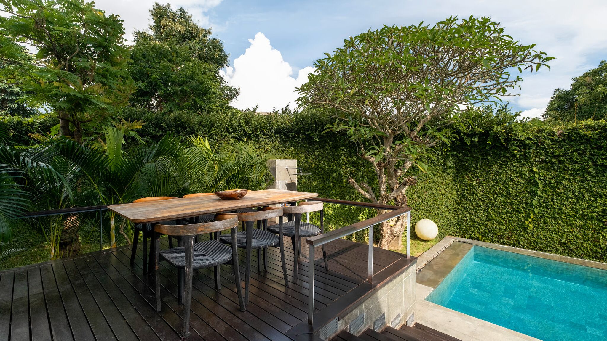 Um deck de madeira com mesa e cadeiras, piscina e vegetação exuberante.