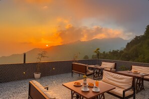 Um terraço com assentos e mesas, uma parede com padrão decorativo e vista do pôr do sol sobre montanhas.
