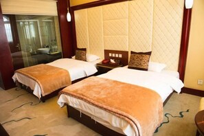 Quarto de hotel com duas camas, um cabeceira acolchoada grande, uma mesa de cabeceira com abajur e vista para o banheiro por uma porta de vidro.
