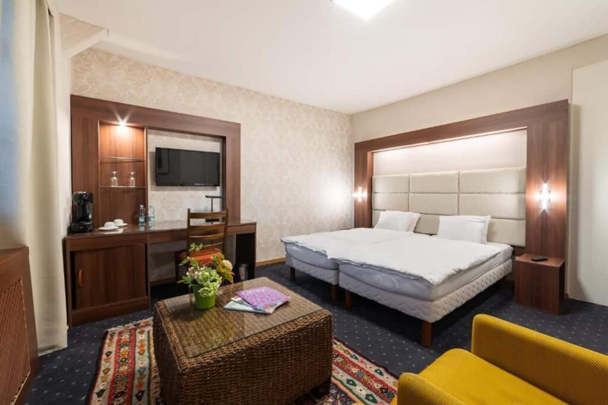 Quarto de hotel com uma cama, uma escrivaninha com uma televisão, uma cadeira, uma pequena mesa com uma planta e uma poltrona amarela.