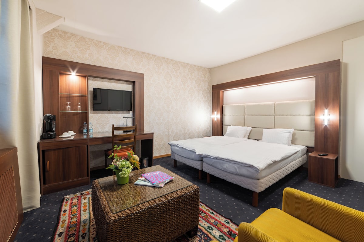 Quarto de hotel com cama, escrivaninha com televisão, mesinha com planta e poltrona amarela.