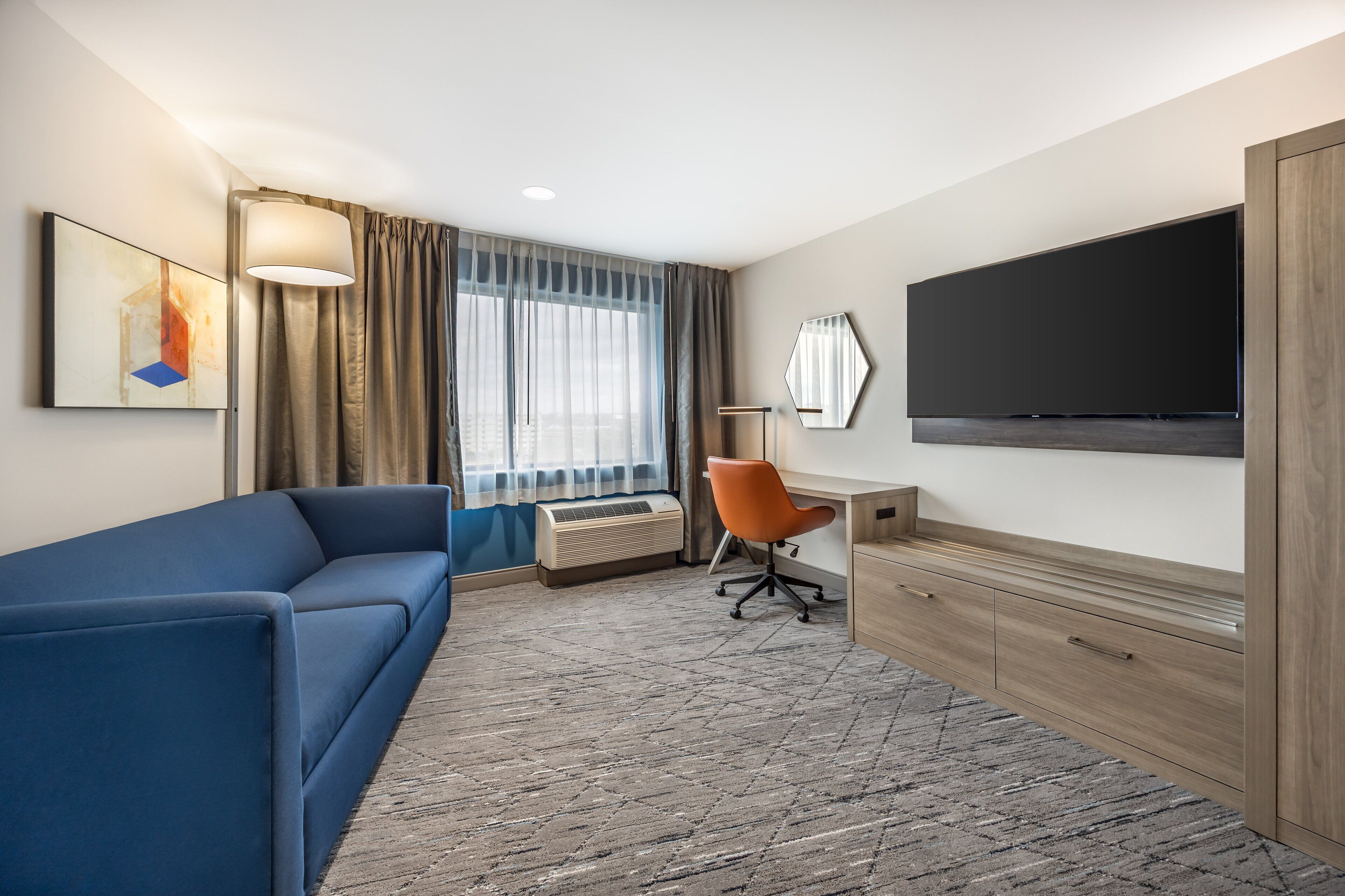 Quarto de hotel com um sofá azul, uma cadeira de escritório laranja, uma televisão de tela plana e uma janela grande com cortinas.