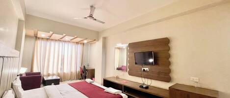 Quarto de hotel com duas camas, televisão fixada em um painel de madeira, ventilador de teto e uma mesa com cadeira.